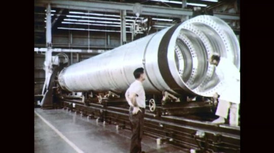 1960s: Men inspect rocket booster tank. Men lift rocket booster tank ...