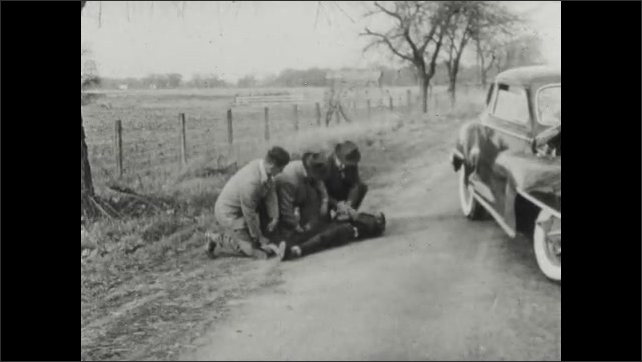 1950s: Three men kneel by injured man lying on side of road next to car ...