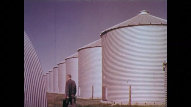 1960s: Field of wheat and grain silos. Man walks by grain silos ...