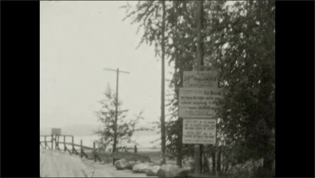 1920s: Gateway with sign. Car drives down road. Signs on roadside next ...