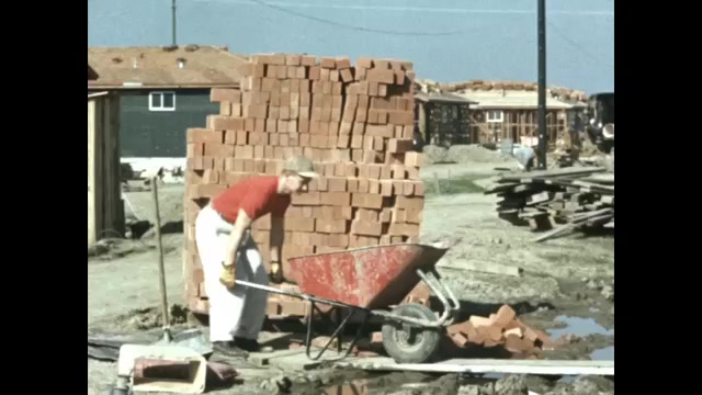 1950s: Boy pushing wheelbarrow with bricks. Woman with clapboard. Man constructing chimney on ...