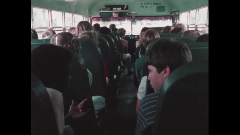 1980s: Children ride in a school bus. Boy hands a book to another boy ...