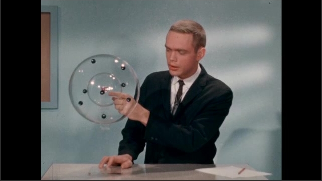 1960s: Man standing with electron model, talking. Man touching model ...