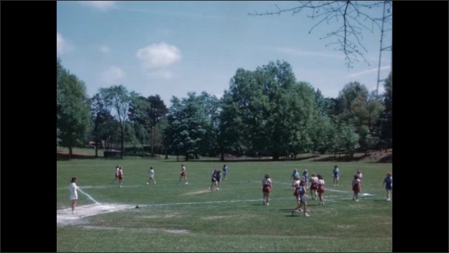 1940s: Girls play game of softball on field. People sit on grass ...