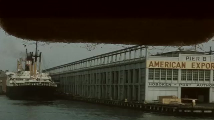 1950s: Cargo ship at dock next to a warehouse on a pier. Tug boat pulls ...