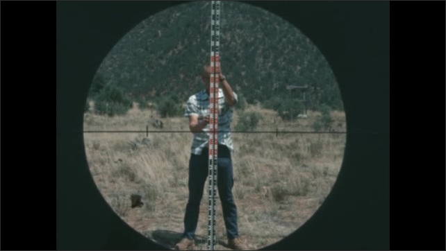 1950s: Scrubby landscape, man stands still in crosshairs, holds pole ...