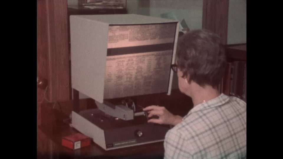 1970s: Woman at a microfiche reader skims through a film. She stops ...
