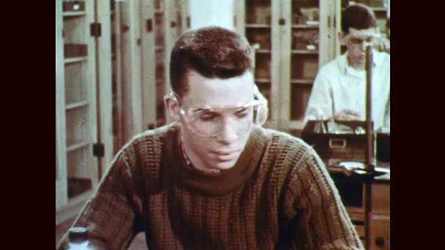1950s: Boy looks to side. Hand holds test tube over flame. Girl turns ...