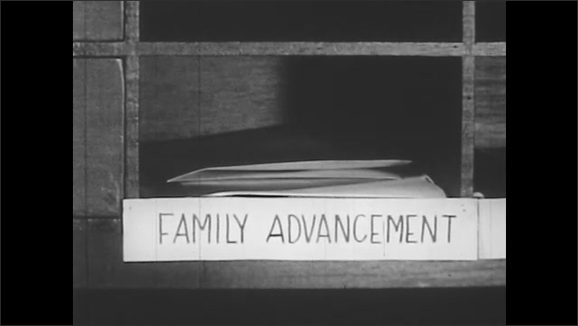 1950s: Desk and drawers. Sign, “Family Advancement.” Hands tear check ...